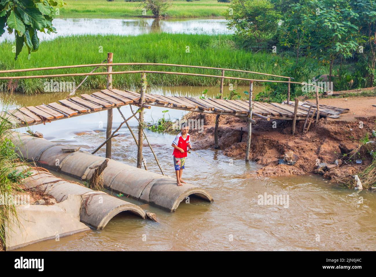 A Thai boy is fishing in a stream surrounded by rice fields and trees ...