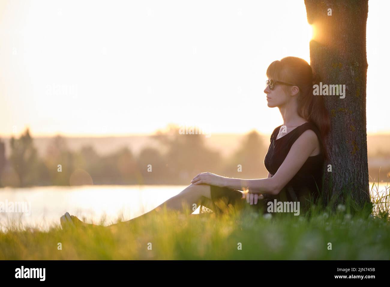 Lonely young woman sitting alone on grass lawn on lake shore enjoying ...