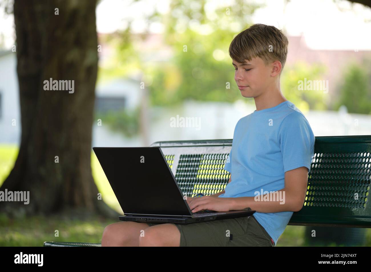 Teenager school boy typing on laptop computer sitting on bench outdoors ...