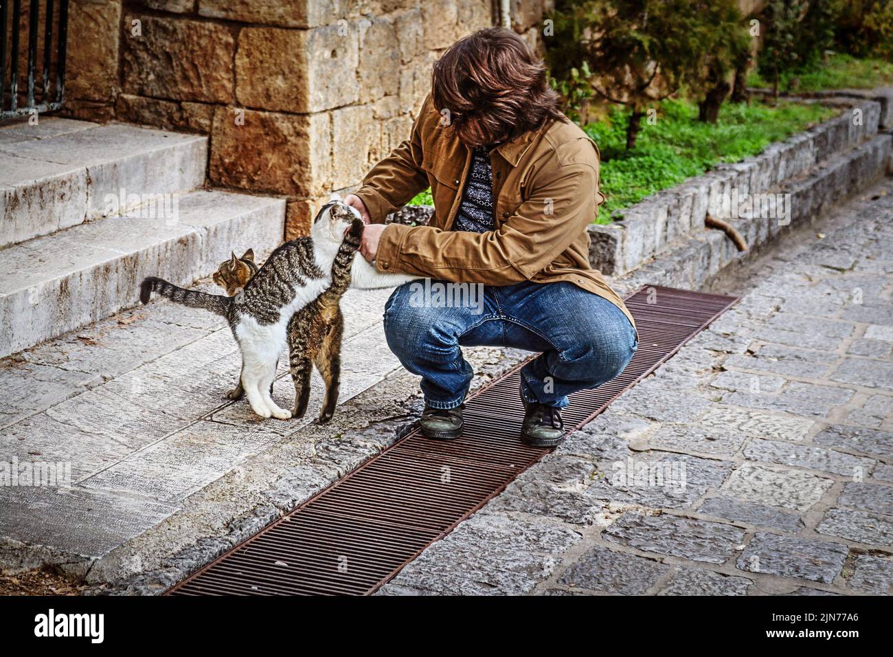 Young man in jeans and jacket with longish hair kneeling down to pet to ...