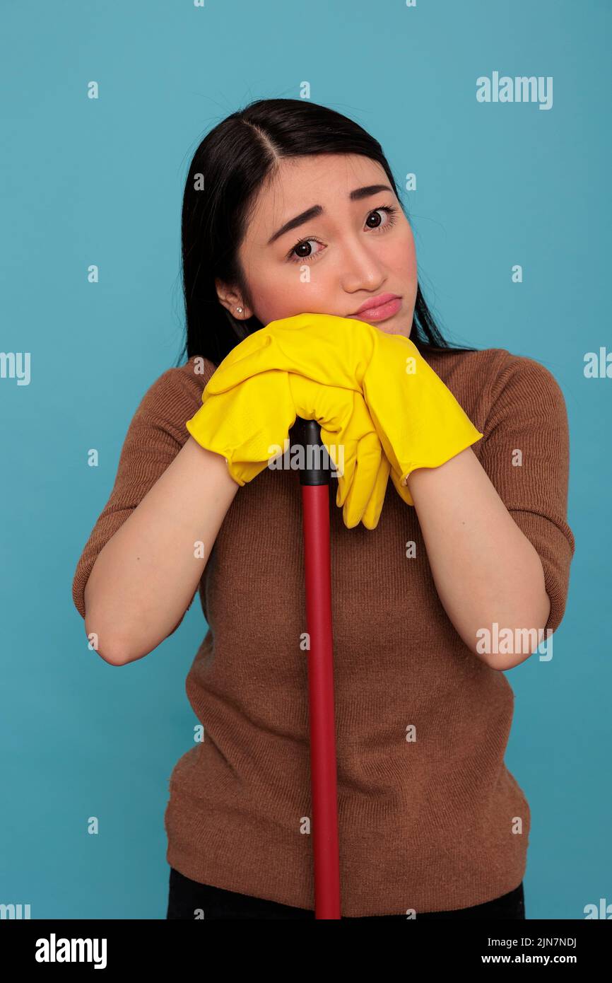 Overwhelmed sad asian housewife holding on brush stick yellow gloves ...