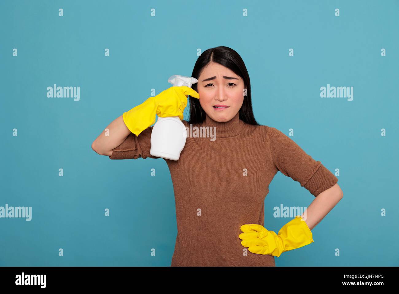 Frustrated tired young asian housemaid holding a spray near her temple ...