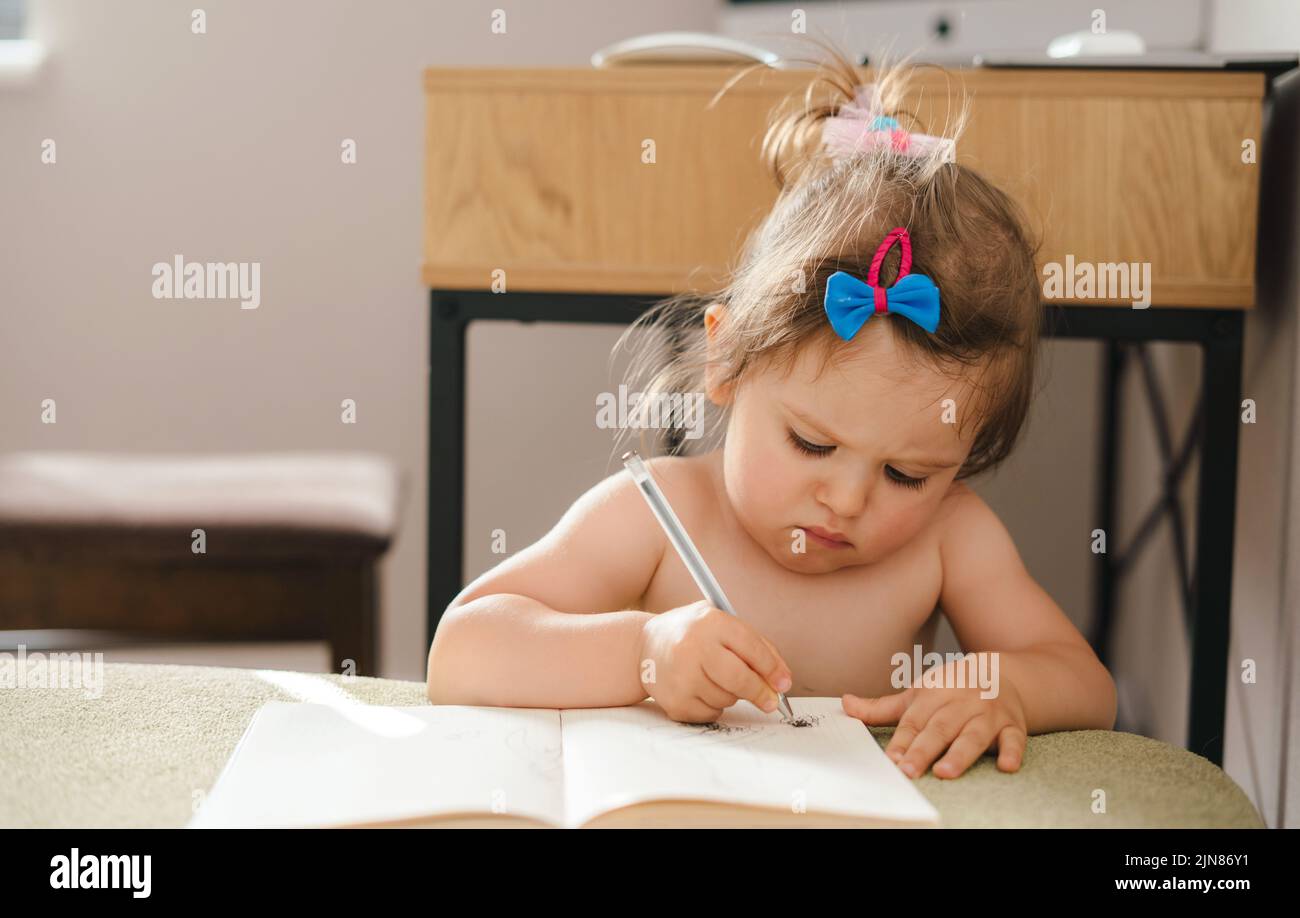 Little baby girl sitting at a table and trying to write letters, as an ...