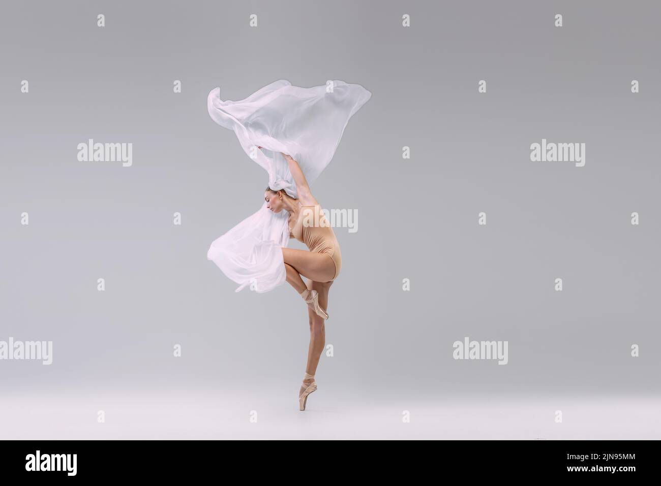 Portrait of young graceful ballerina dancing with fabric isolated over grey studio background ...