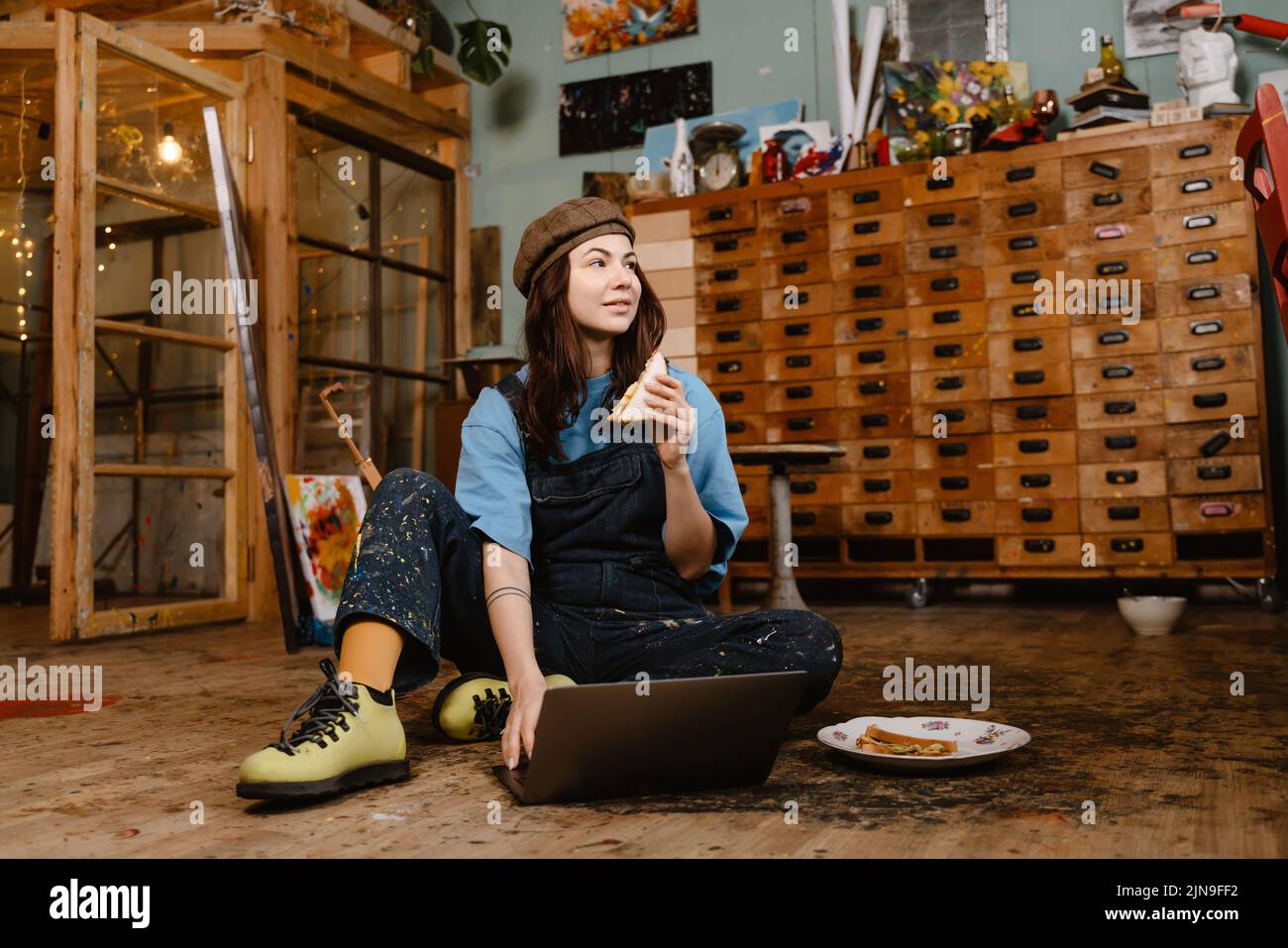 Young woman artist eating sandwich while using laptop in her studio ...