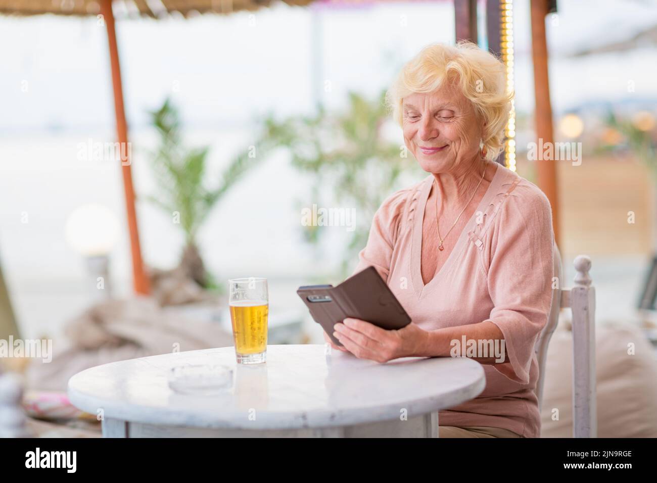 Mature woman sitting at a table in a summer cafe and drinking beer ...