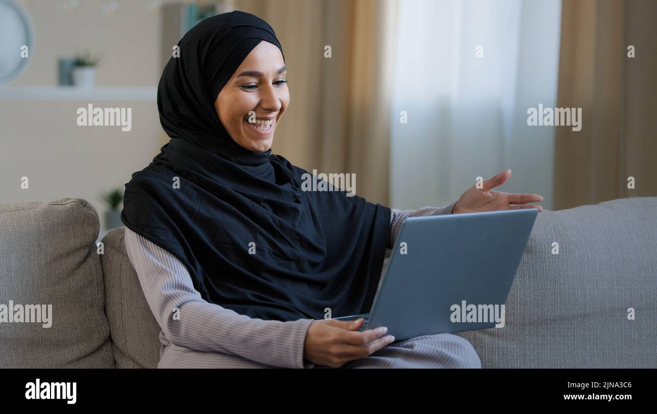 Friendly Arabian Muslim Woman Girl In Hijab Sit On Cozy Sofa Talk Video Call With Friend