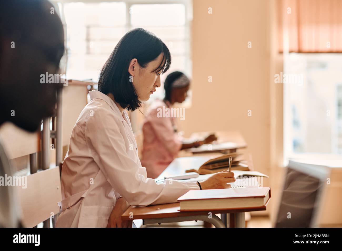 Asian medical student in white coat writing test at desk during lecture ...