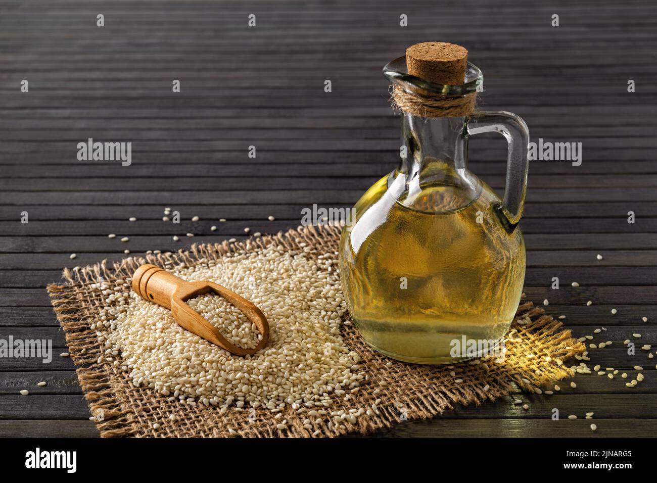 Fresh sesame oil in a glass jug and heap of white til seeds over black ...