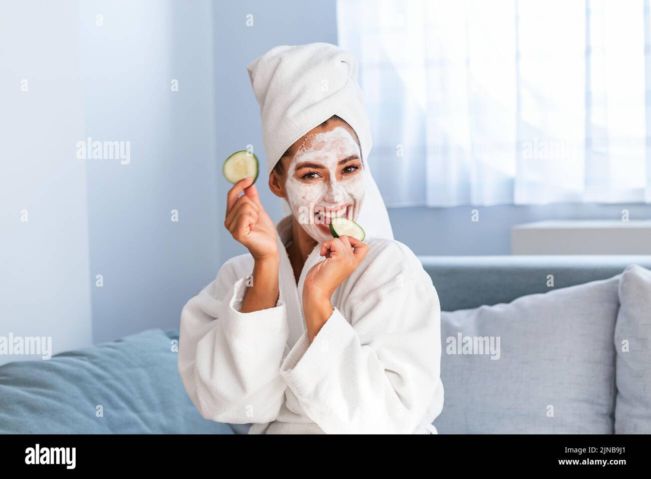 Beautiful young woman with facial mask on her face holding slices of fresh cucumber. Skin care ...