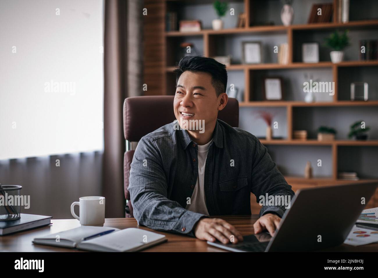 Happy mature chinese man at table working with laptop in home office ...