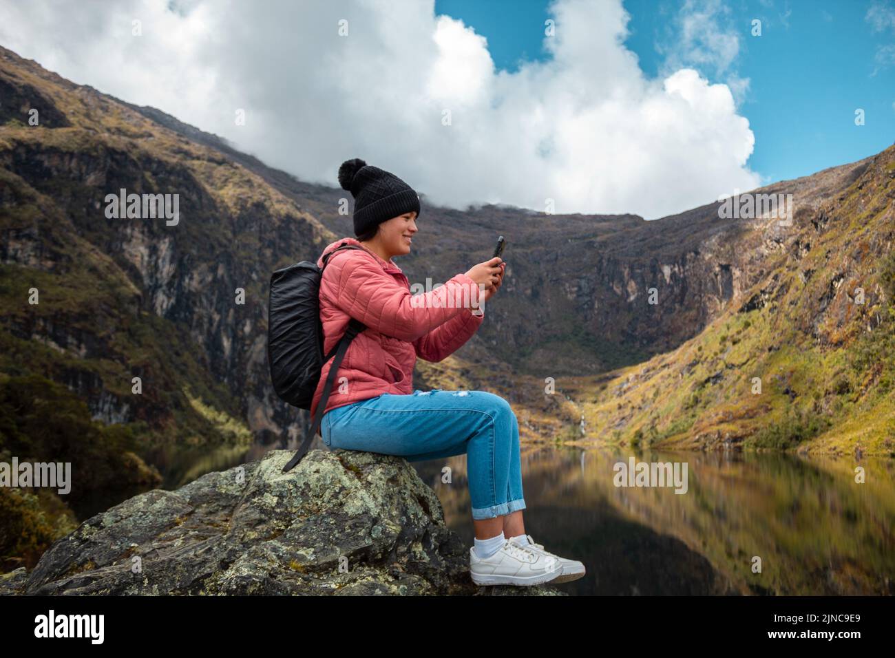 An attractive hiker sitting and holding a phone next to a tranquil lake ...