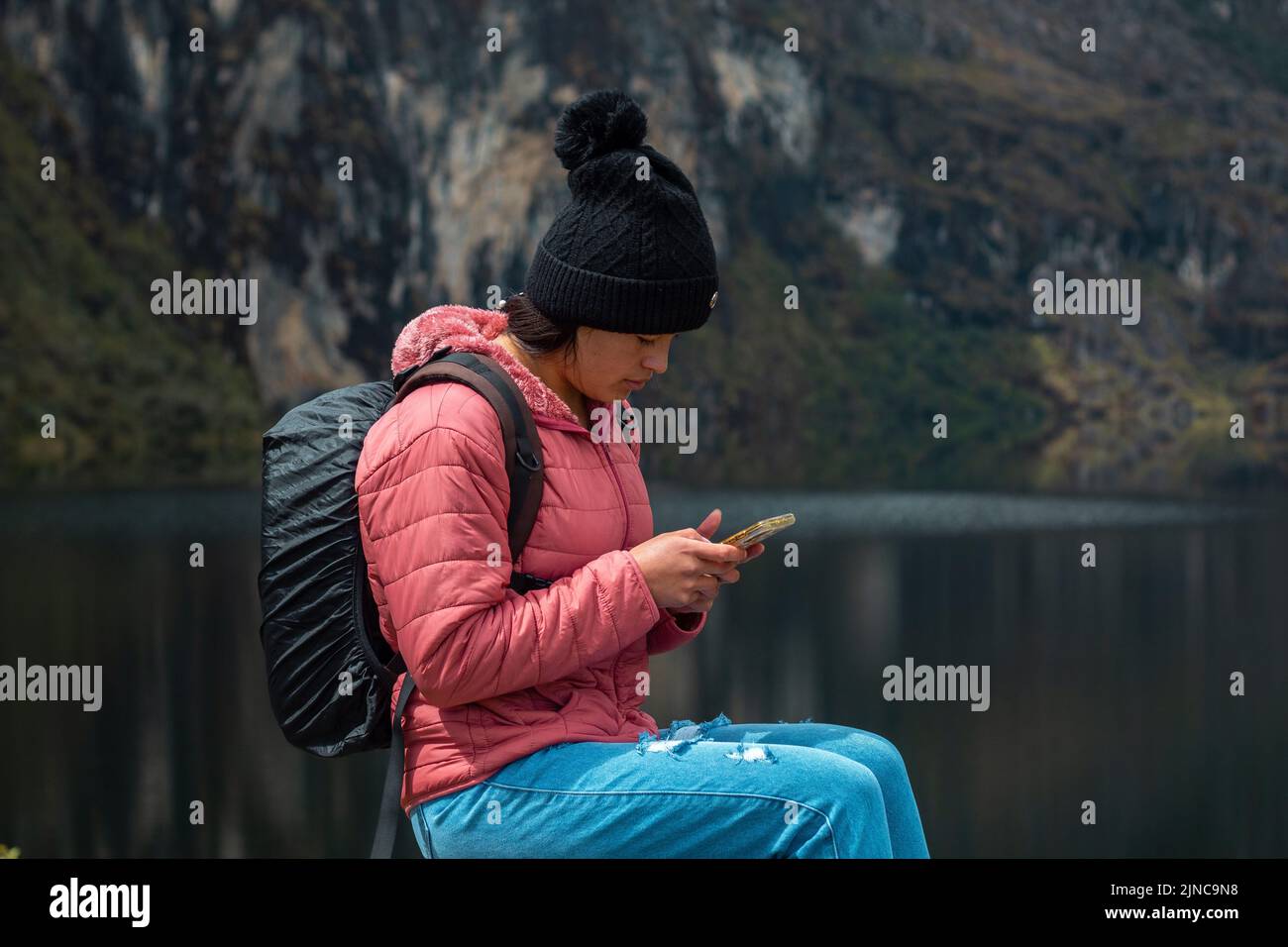 An attractive hiker sitting and holding a phone next to a tranquil lake ...