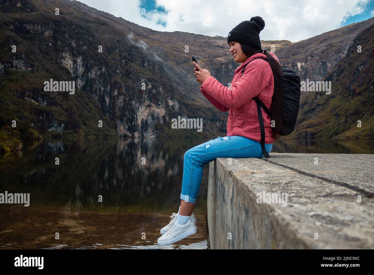 An attractive hiker sitting and holding a phone next to a tranquil lake ...