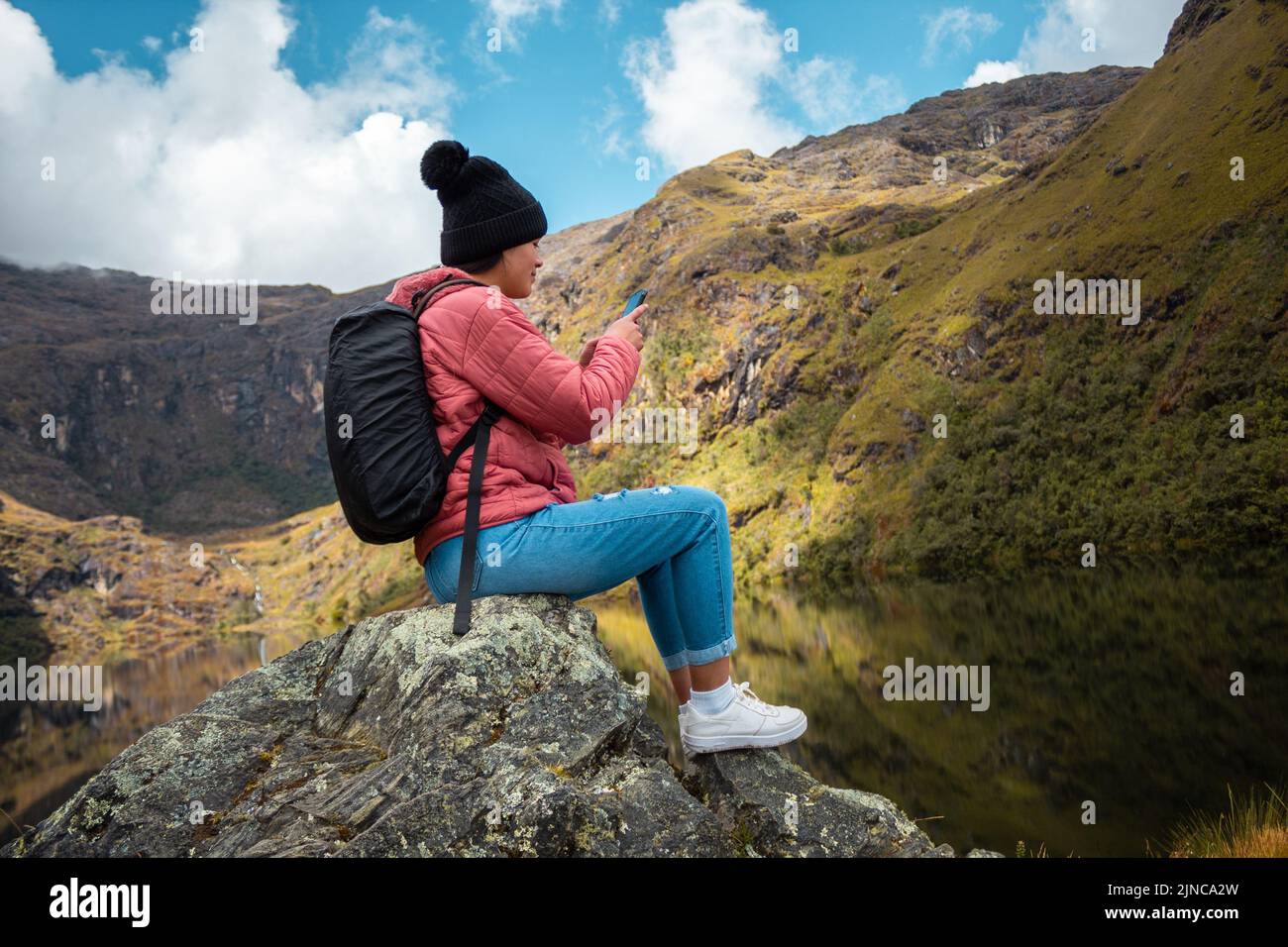 An attractive hiker sitting and holding a phone next to a tranquil lake ...