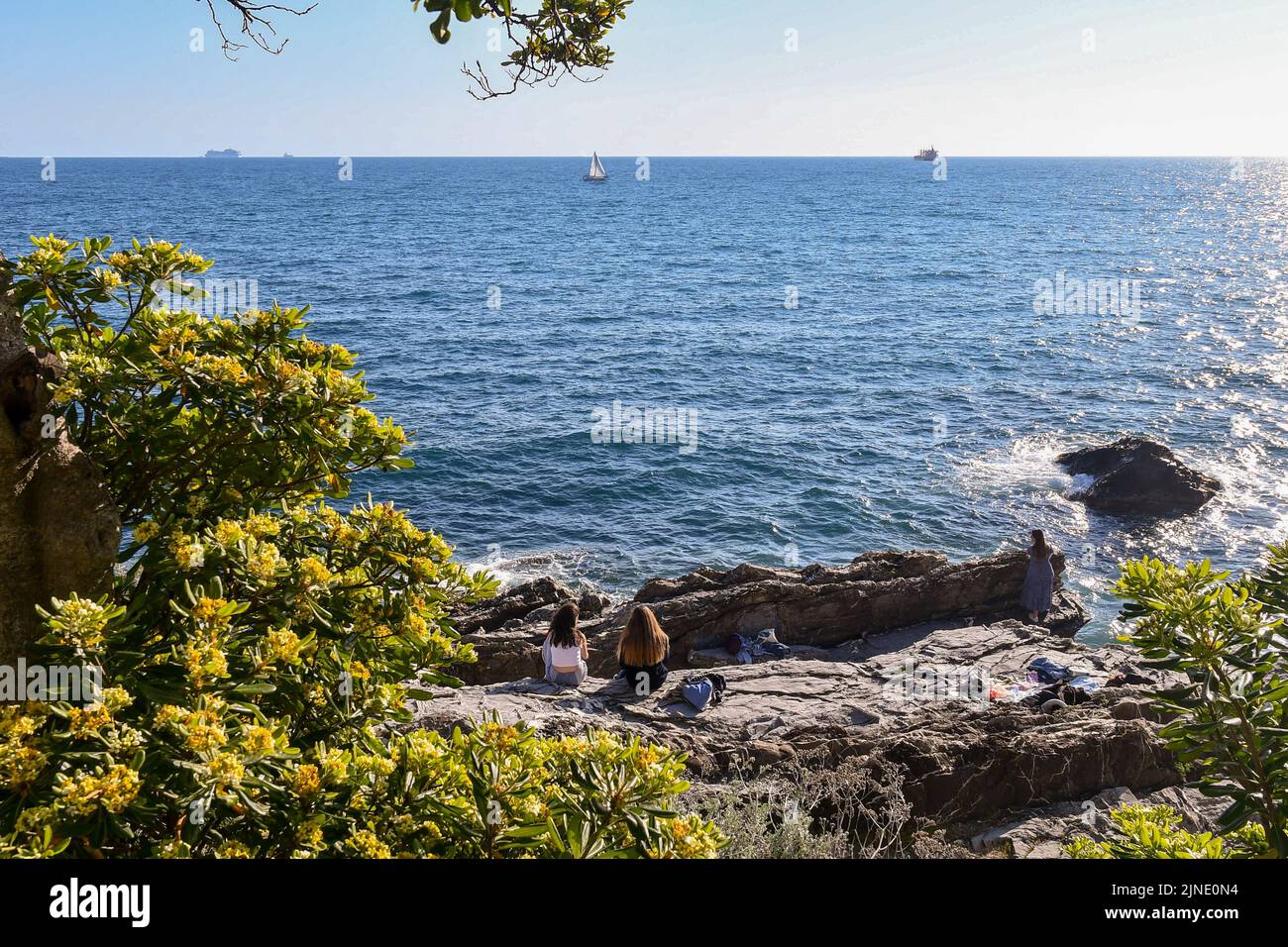 Seascape view from the promenade with girls sitting on the rocks by the sea and flowering plants ...