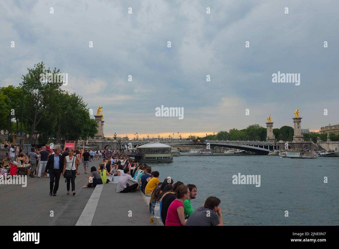 People, mostly tourists, sit and relax by the Seine river, near the ornate bridge, Pont ...