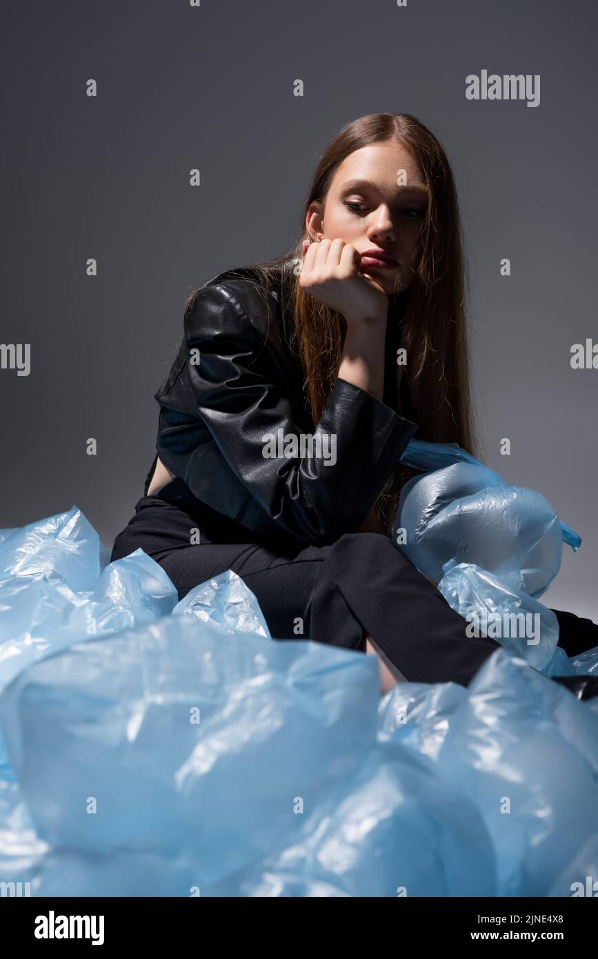 sad model in stylish black suit posing around blue plastic bags on grey ...
