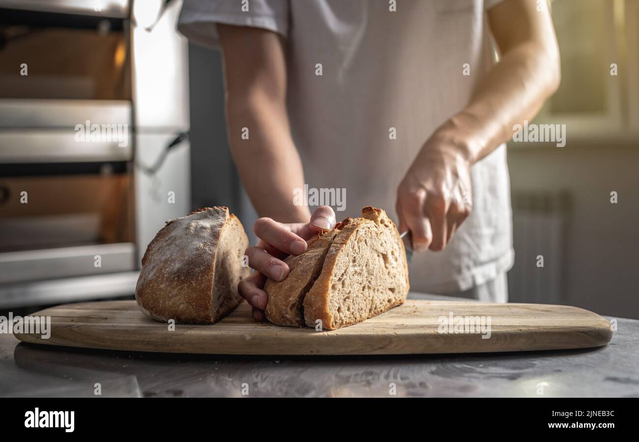 A male baker cuts freshly baked bread with a knife to check the quality ...