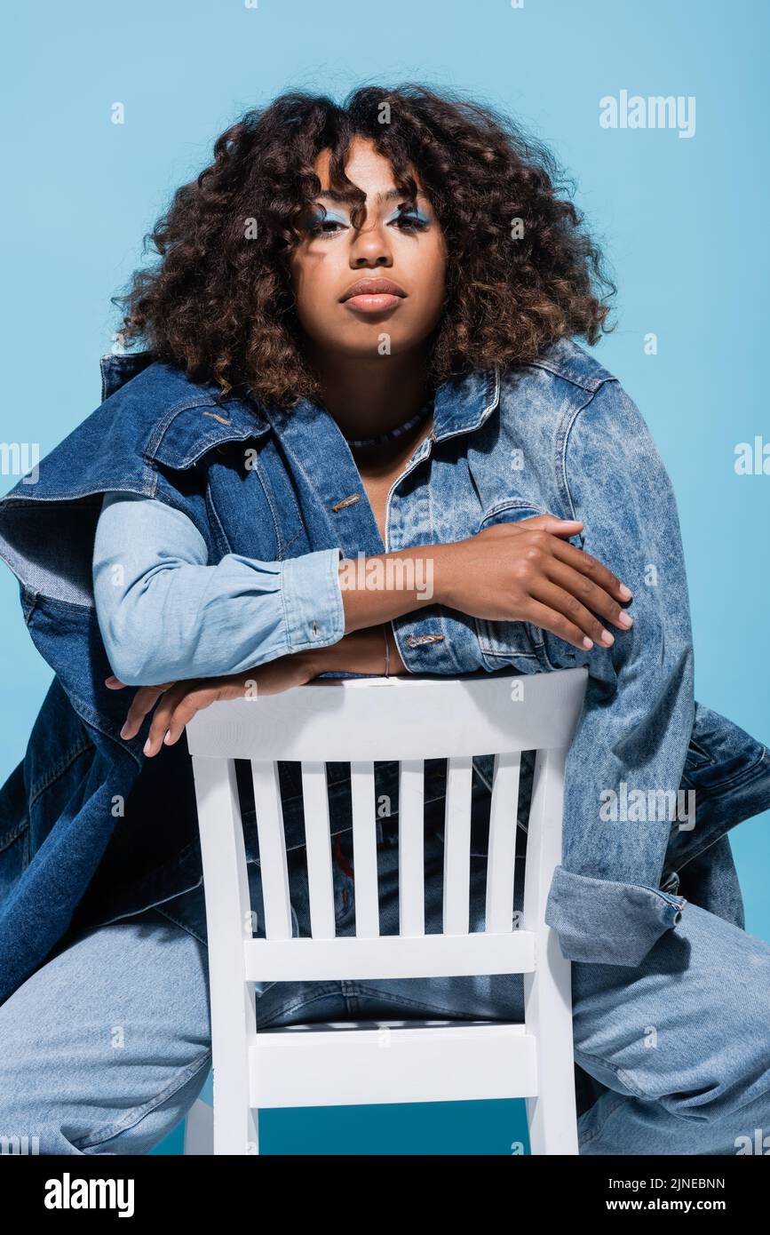 stylish african american woman with wavy hair sitting on chair with crossed arms isolated on ...