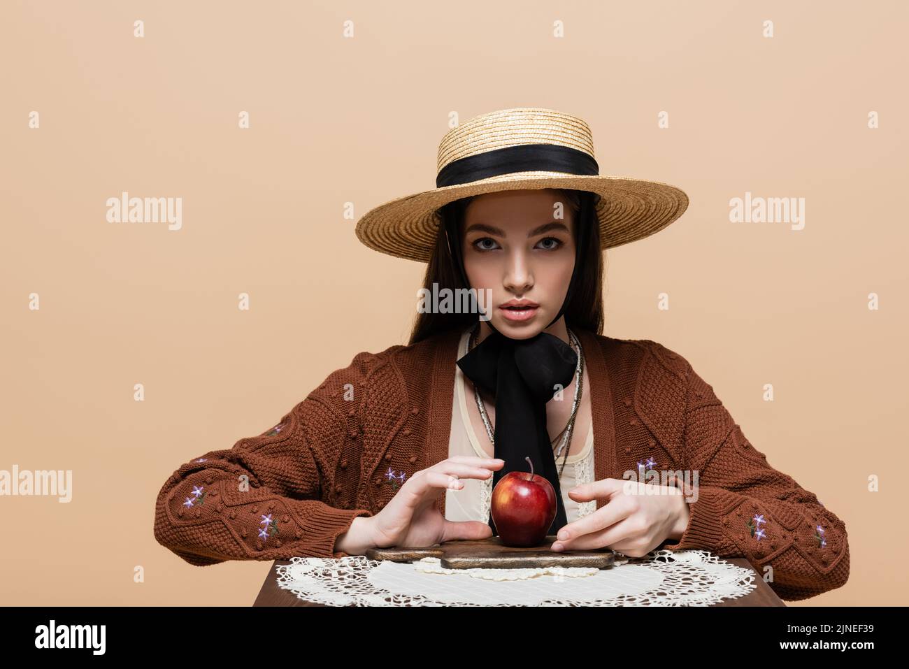 Brunette model in straw hat touching apple on table isolated on beige ...