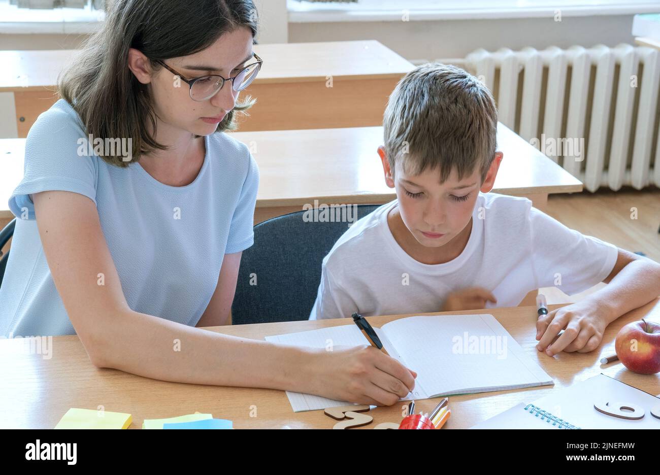 A boy and a teacher are sitting at a table with notebooks and pens. A ...