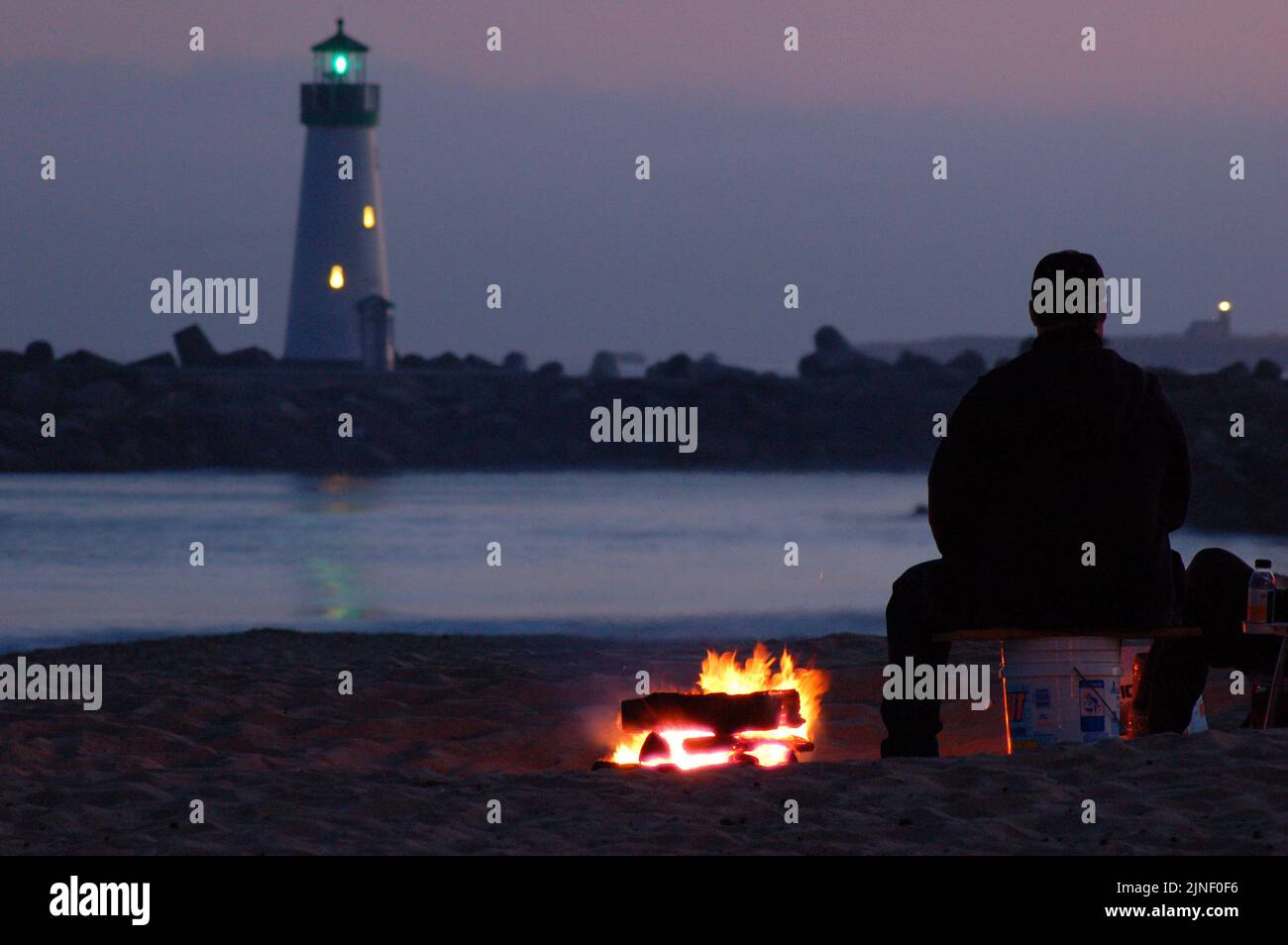 An adult man sits by a bonfire at within sight of a lighthouse on the ...