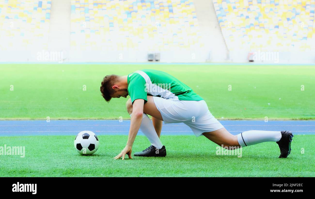 young football player in uniform stretching legs on green grass bear ...