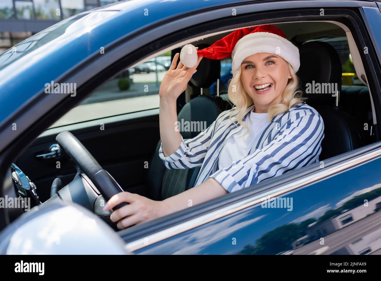 Positive blonde driver in santa hat looking at camera while driving car ...