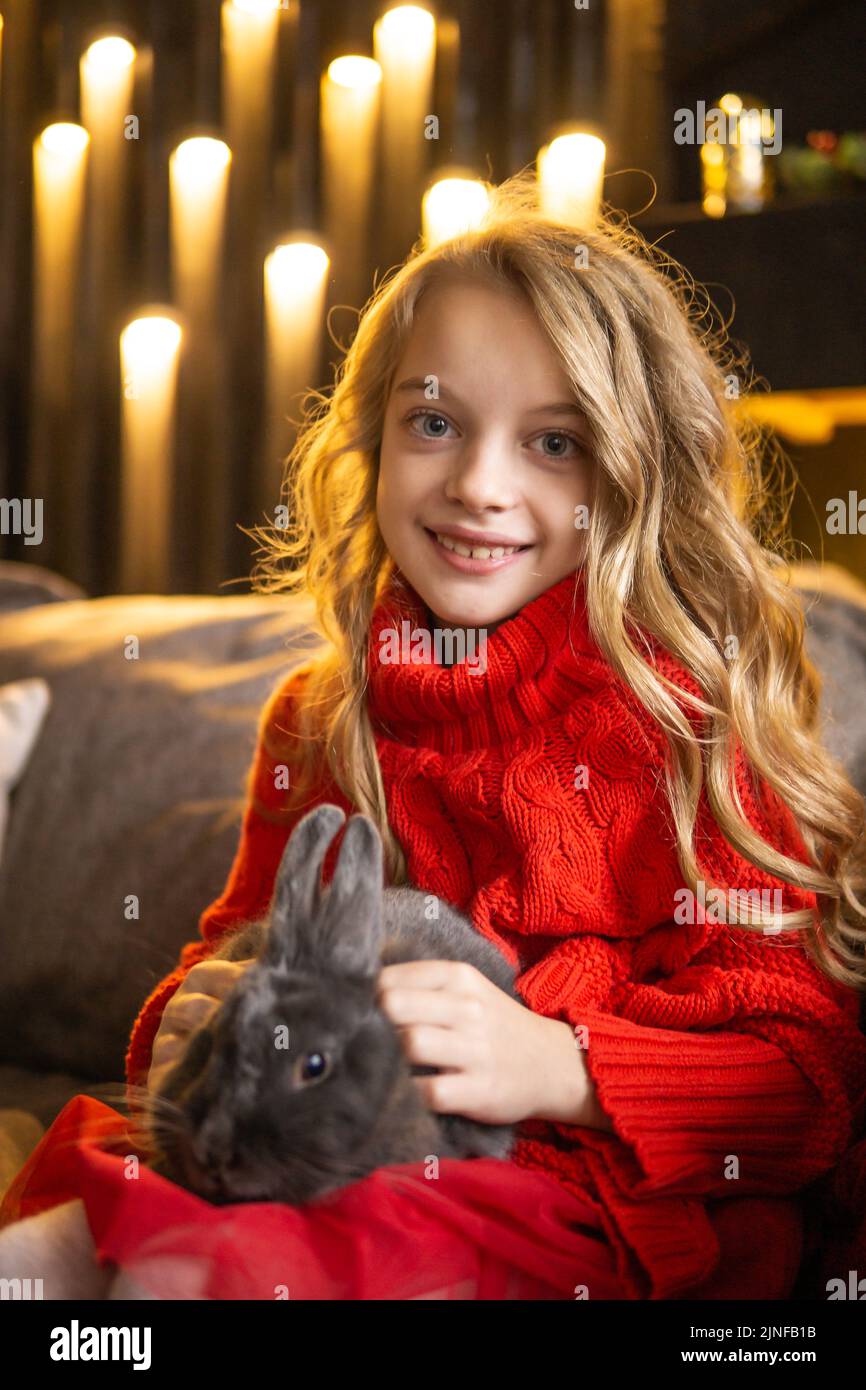 A little blonde girl with a gray rabbit in her arms next to fireplace ...