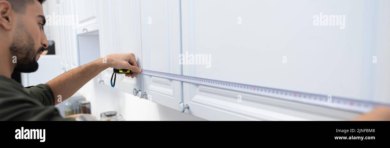 Side view of arabian man measuring cupboards in kitchen at home, banner ...