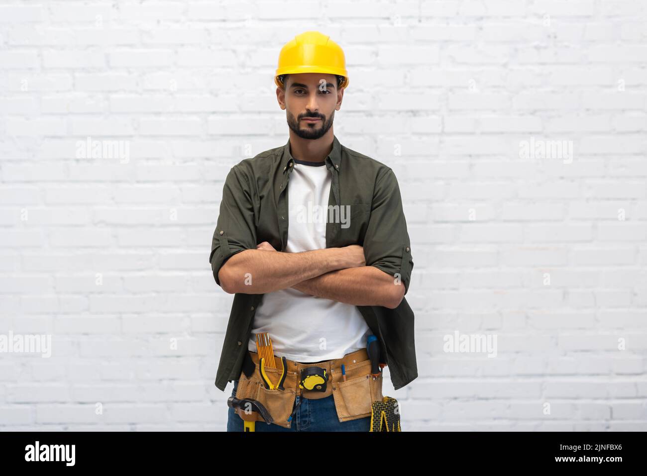 Young muslim handyman in helmet and tool belt looking at camera at home ...