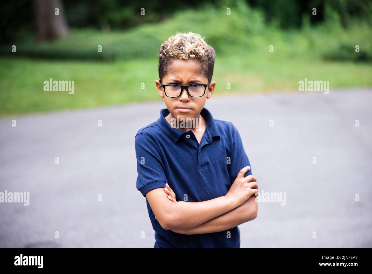 An elementary aged boy with glasses on and his arms crossed with a mad or mean look on his face ...