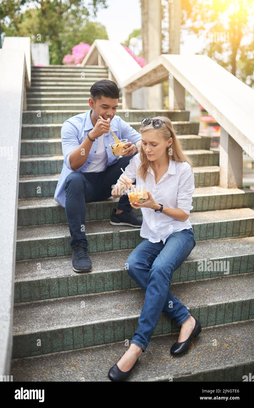 Diverse couple sitting on steps and eating chinese noodles with sticks ...
