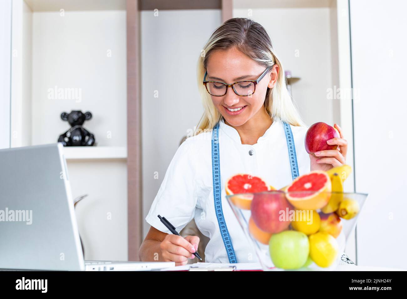 Smiling nutritionist in her office, she is holding a fruit and showing ...