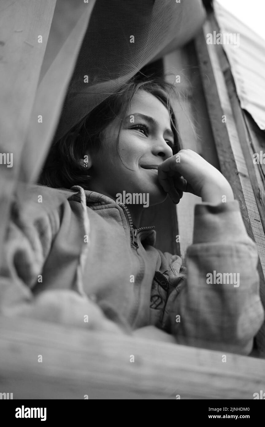 A vertical grayscale shot of a smiling little refugee girl in a camp in ...