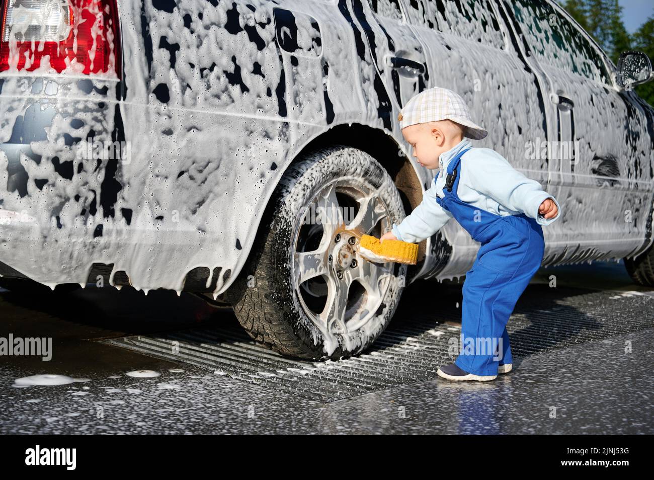 Toddler child leaning over rear wheel of foamy car and holding sponge ...