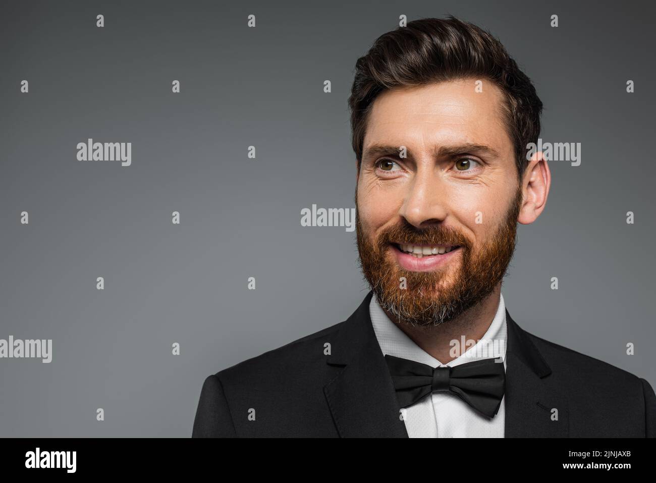 cheerful and bearded man in black tuxedo with bow tie isolated on grey ...
