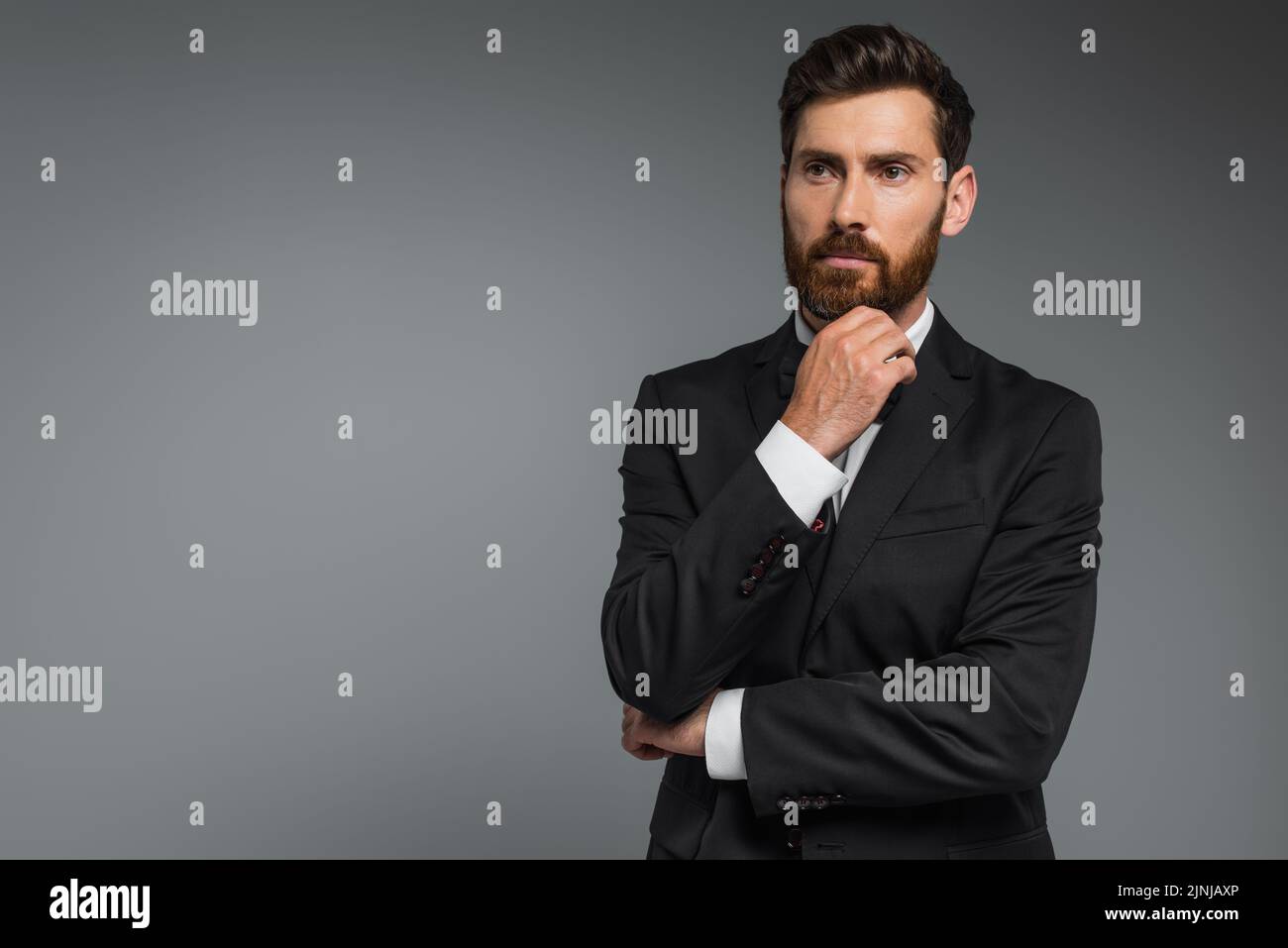 pensive man with beard standing in elegant suit with bow tie looking ...