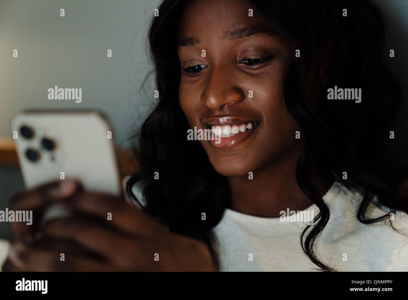 Black Young Woman Smiling And Using Mobile Phone While Lying In Bed At