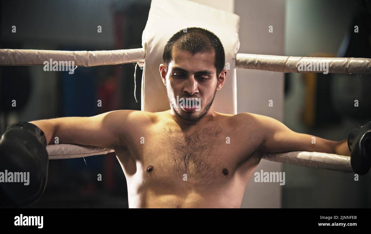 A tired man boxer sitting on the ring in the corner with a teeth guard ...