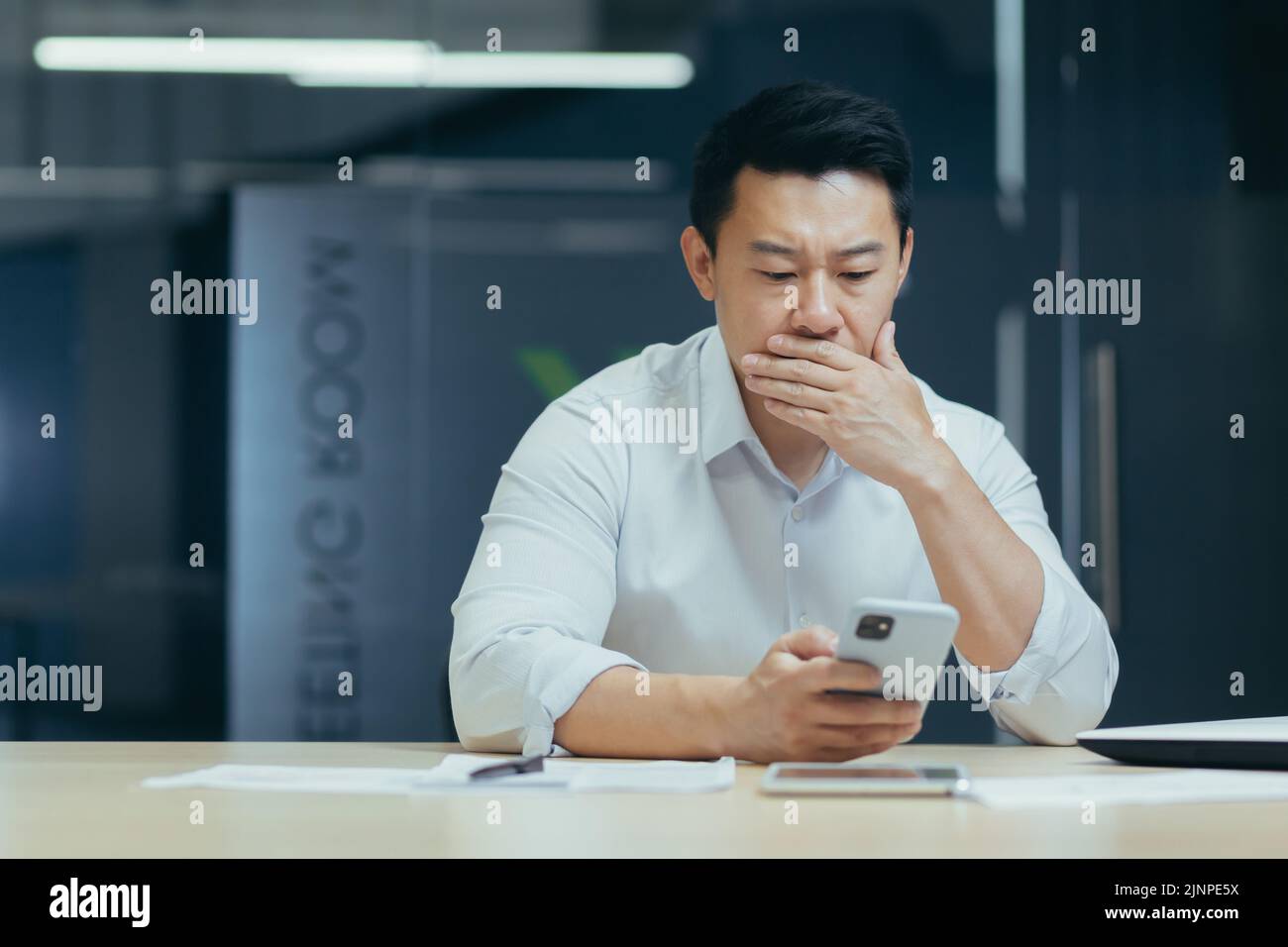 Confused Asian guy holding smart phone feels concerned thinking over ...