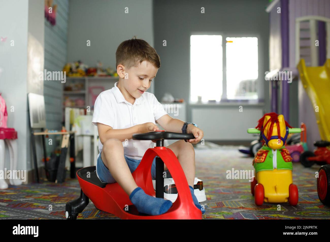 A little preschool boy is riding on a toy white car in the playroom ...