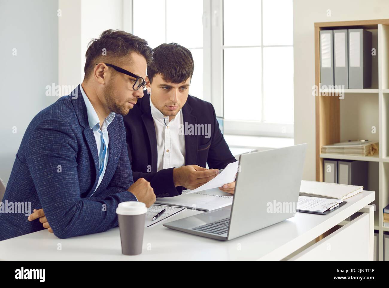 Two serious young men sitting at an office desk and looking at their ...