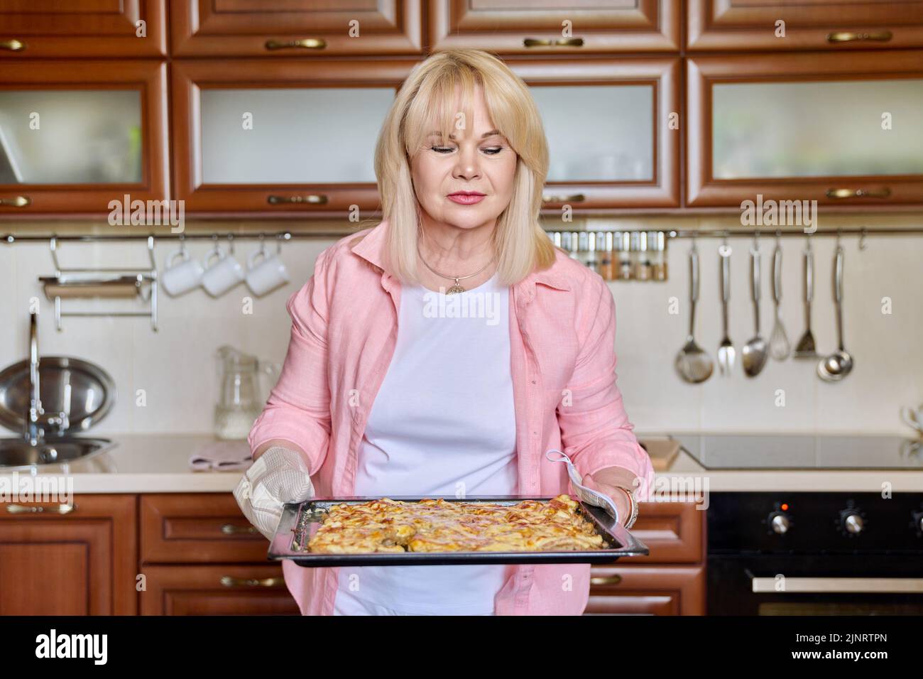 Portrait of mature 50s woman with pan of cooked meat at home in kitchen ...