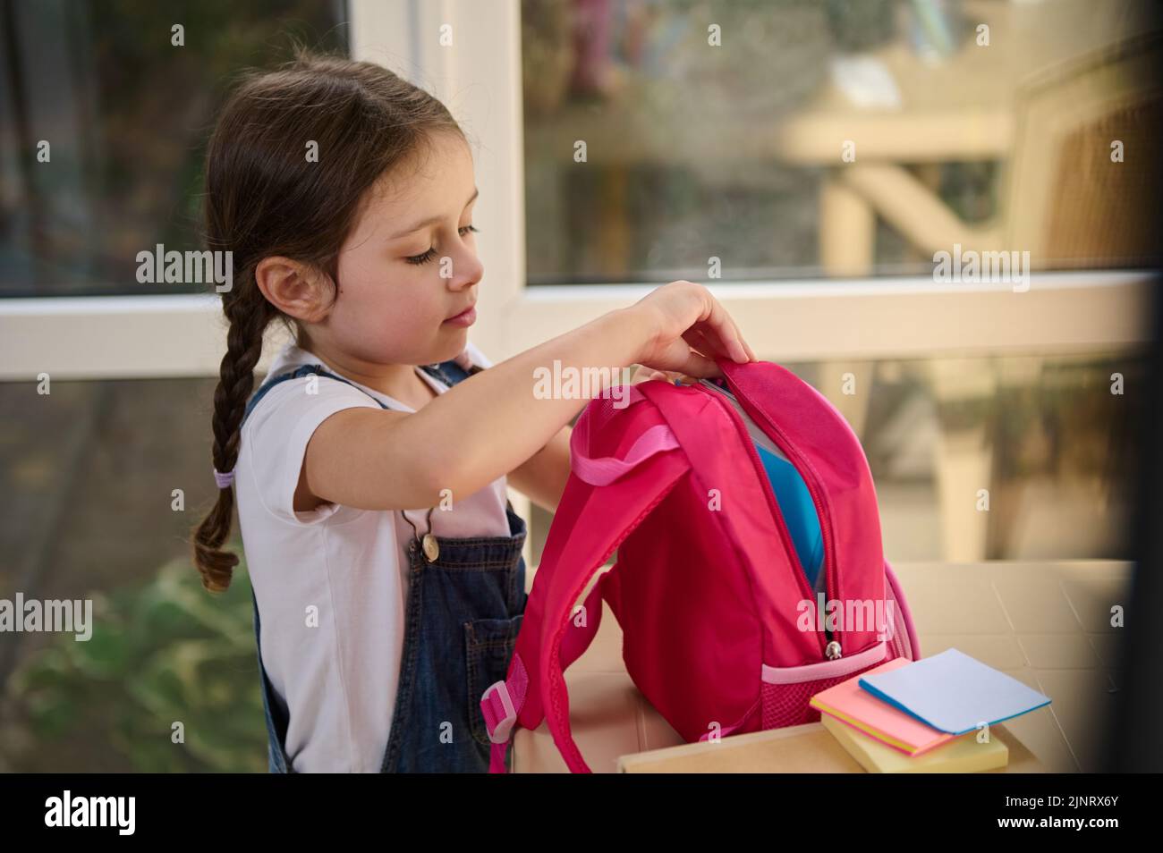 Adorable European little girl puts a notebook in a pink backpack, getting ready for the first ...