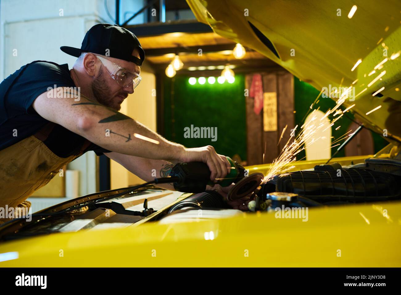 Young repairman bending over engine of car while using electric grinder ...
