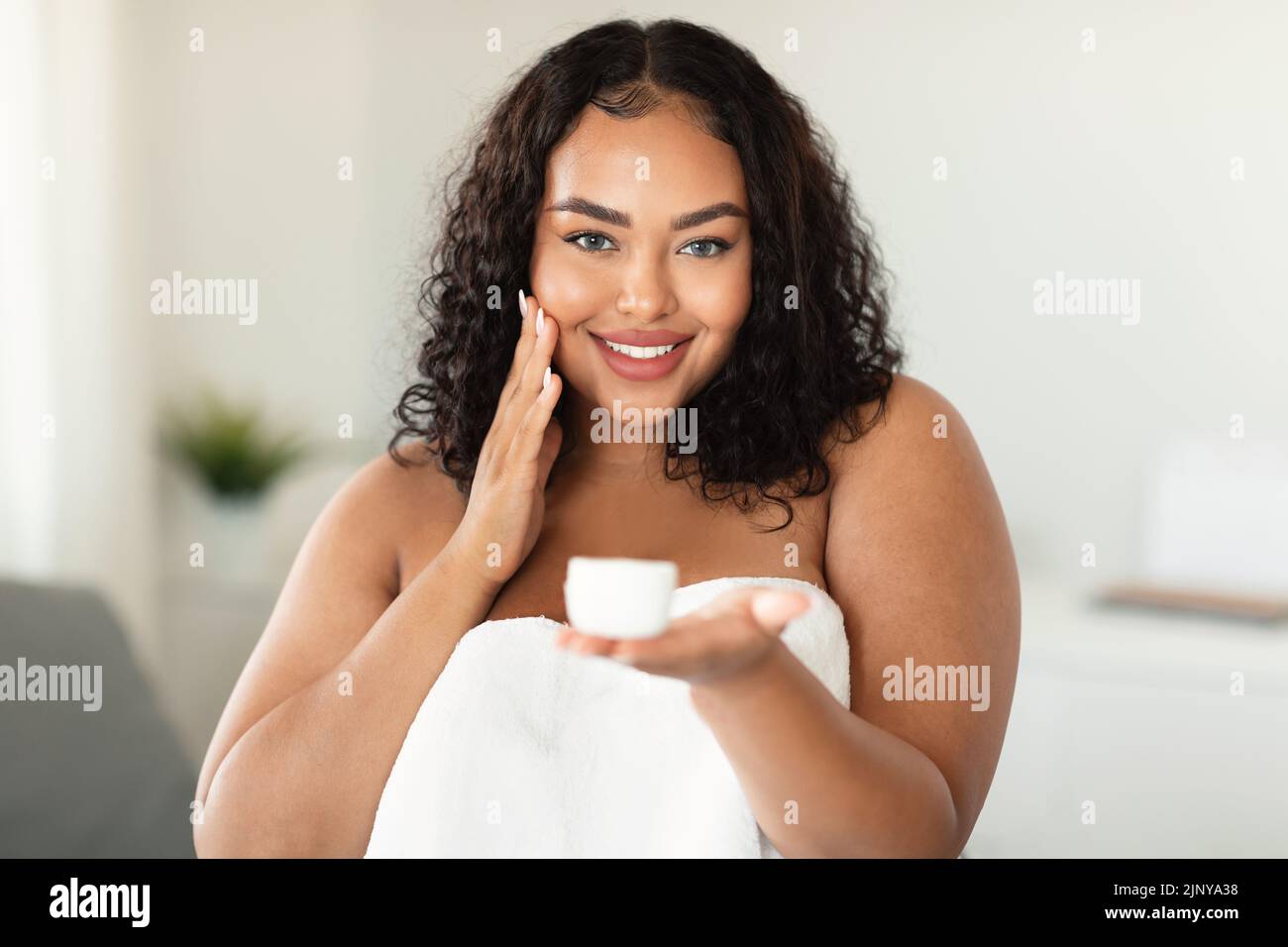 Happy black chubby lady using face cream at home, looking at camera and ...