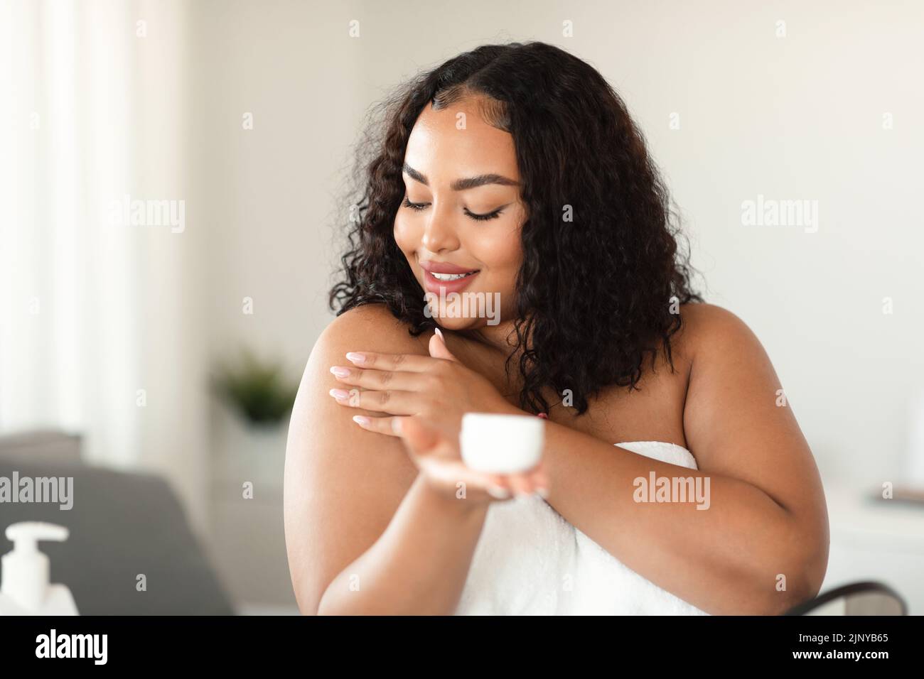 Black plus size lady applying body cream on shoulder after shower in ...