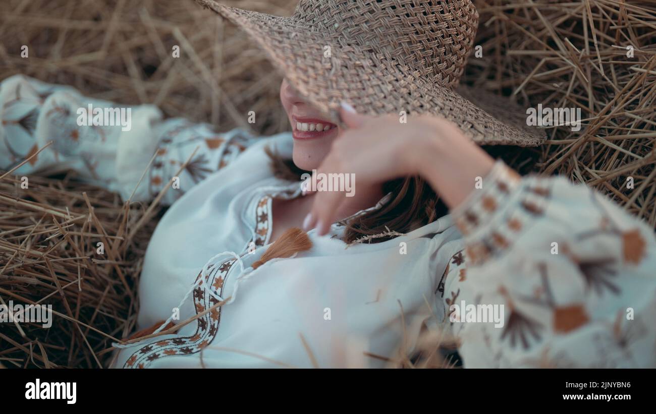 Pretty woman in straw hat and embroidered blouse smiling lying on hay ...